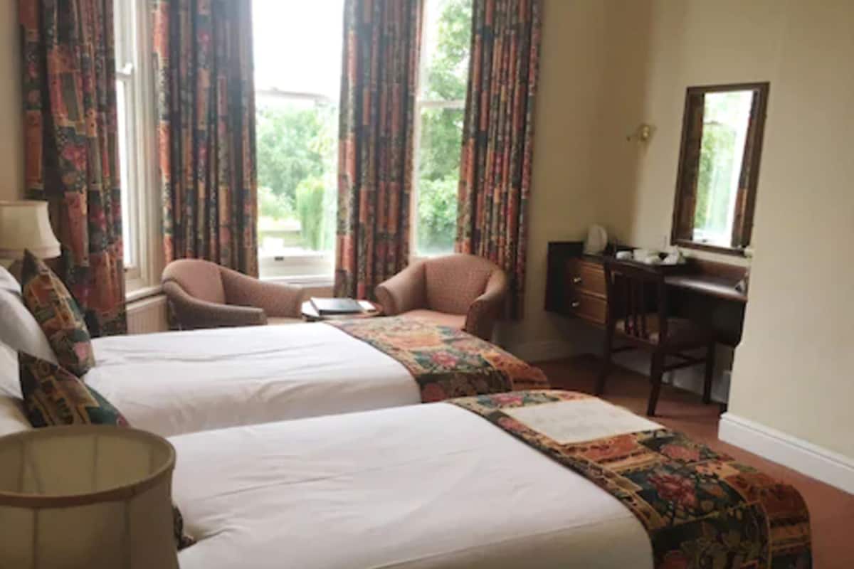 Quarto de hotel com duas camas, uma escrivaninha, uma cadeira e uma janela grande com cortinas.