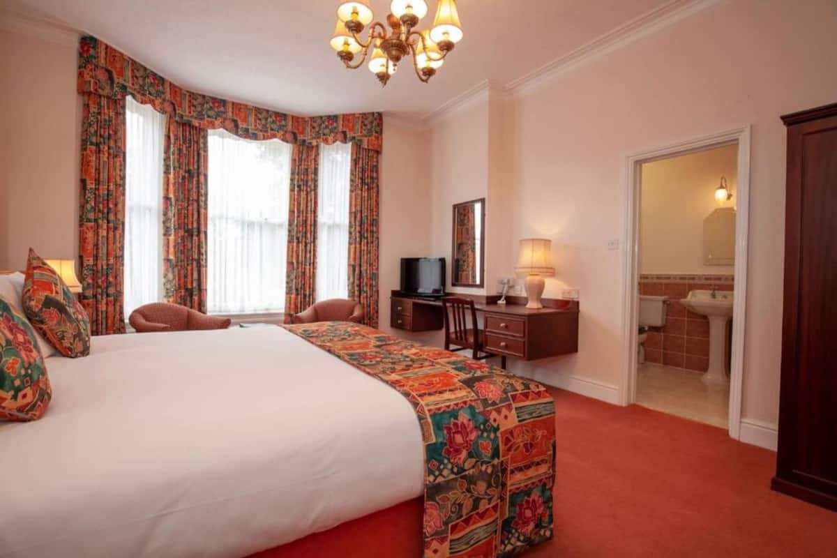 Quarto de hotel com uma cama grande, uma escrivaninha, televisão e banheiro visível por uma porta aberta.