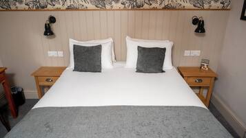 Uma cama bem arrumada com duas almofadas, uma mesa de cabeceira com abajur e uma parede com papel de parede floral.