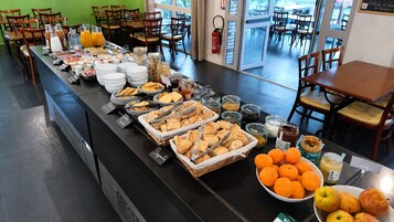 Buffet com diversas opções de comida e bebidas, incluindo frutas e doces, em área de refeições com mesas e cadeiras.