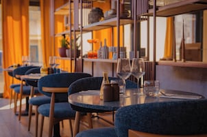Interior de restaurante com mesas redondas, cadeiras azuis e copos de vinho.