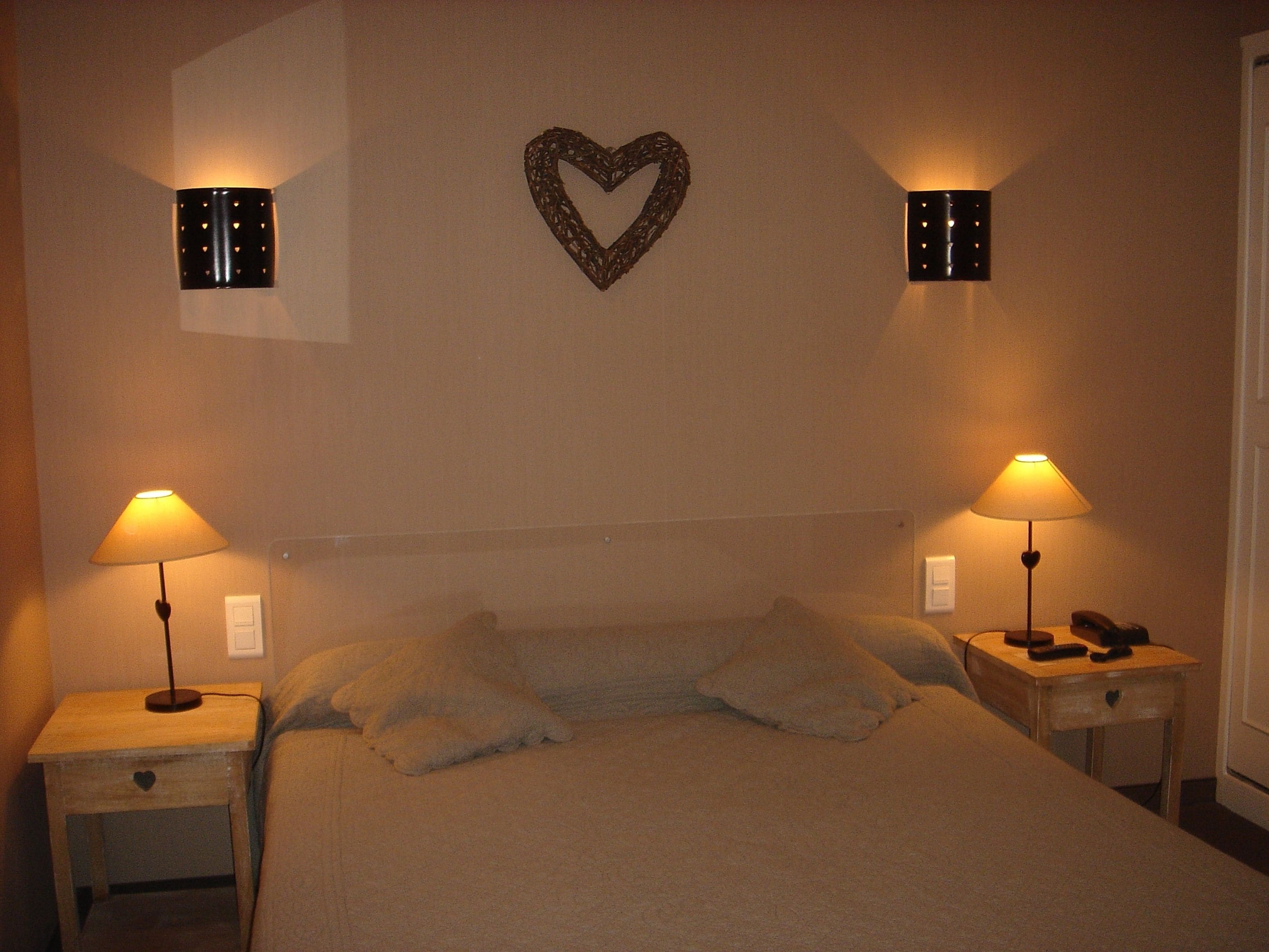 Quarto com cama, duas mesas de cabeceira, duas luminárias fixadas na parede e uma decoração em formato de coração.