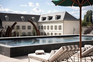 Piscina com espreguiçadeiras e guarda-sol, cercada por um edifício com telhado telhado e várias janelas.