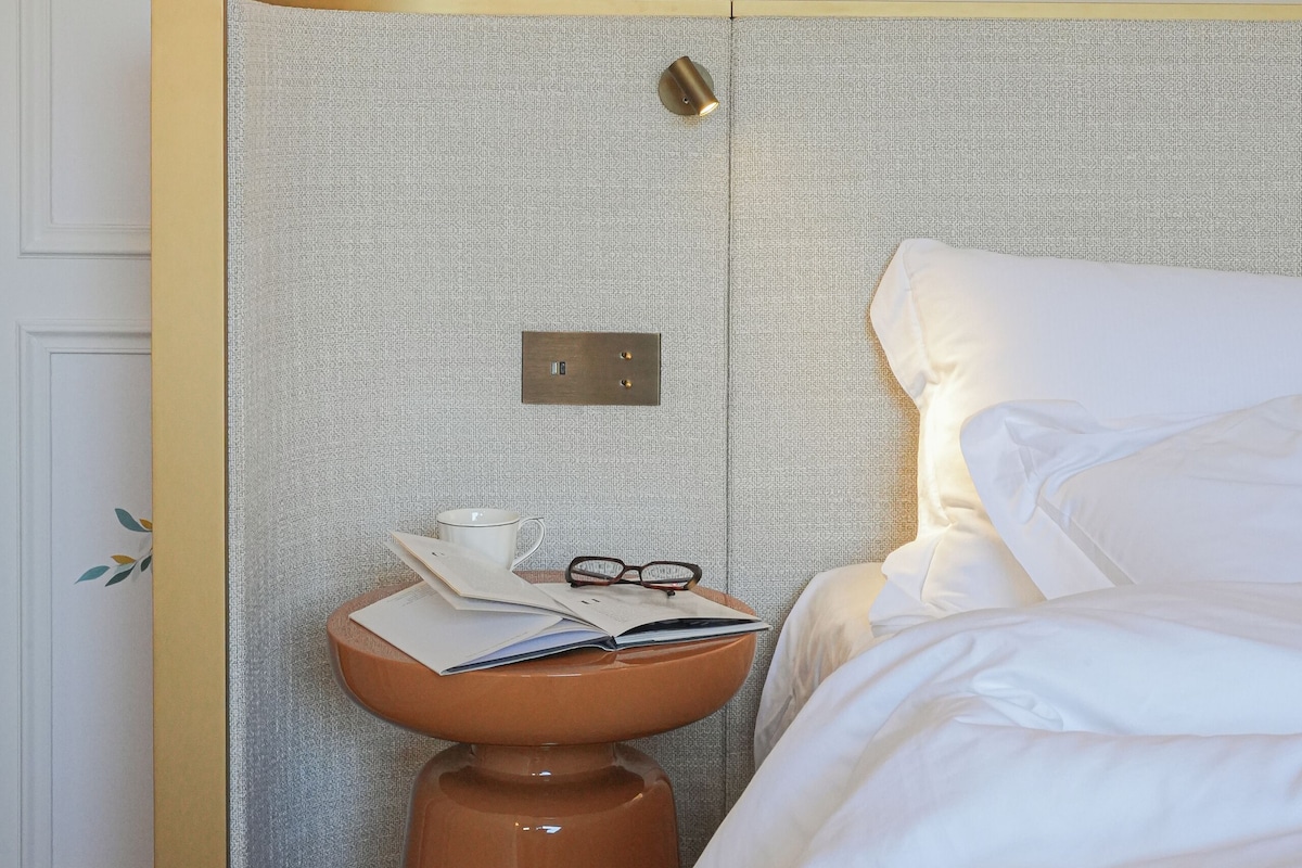 Cama bem arrumada com lençóis brancos, mesa de cabeceira com xícara e copos, e luminária de parede com padrão de folha.