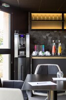 Quarto de hotel compacto com cozinha compacta, geladeira, cafeteira e pequeno bar com copos e lousa de cardápio.