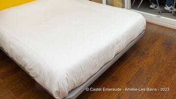 Quarto com cama, guarda-roupa, mesa pequena e varanda com uma pessoa sentada.