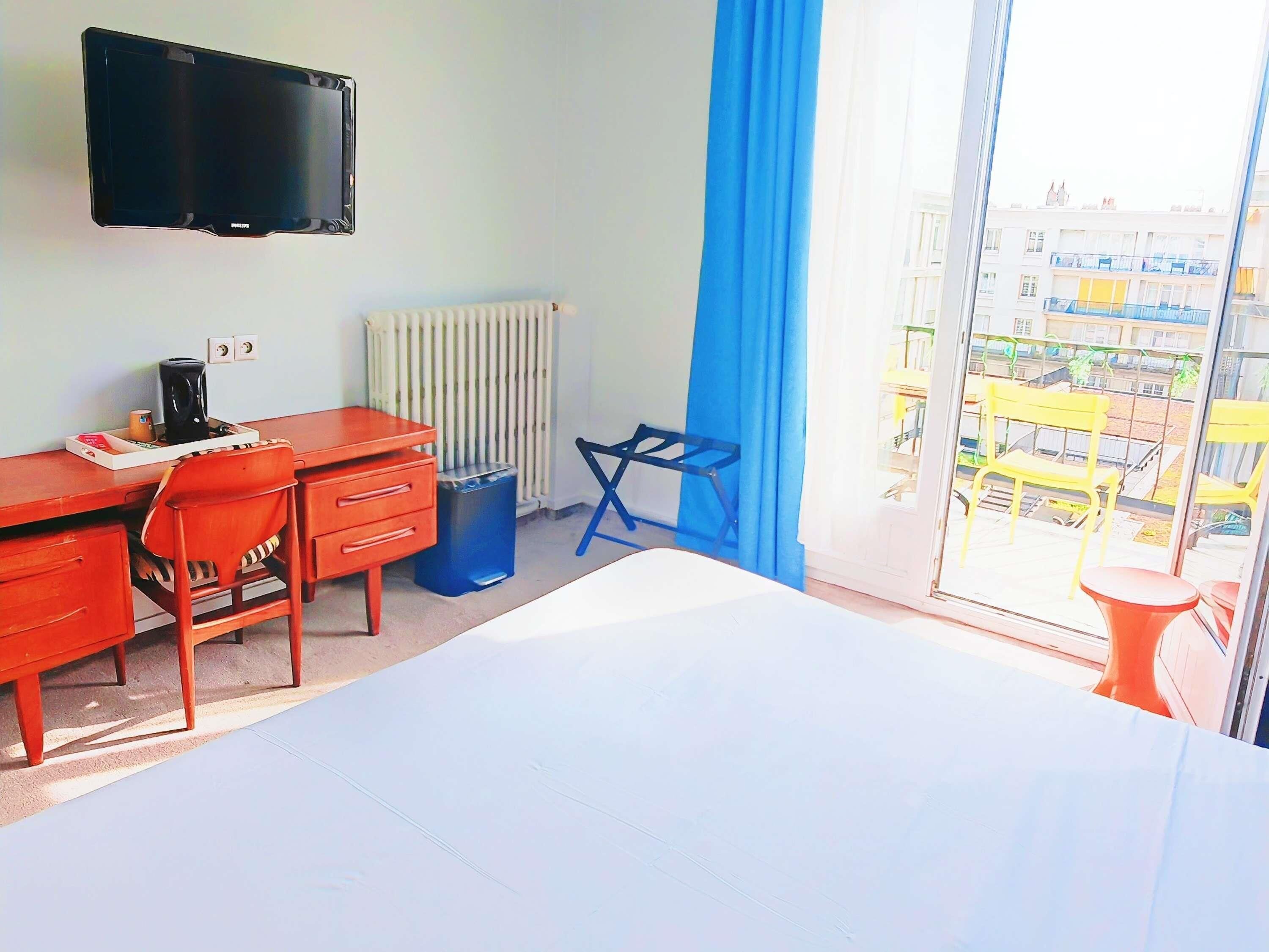 Quarto de hotel com cama, secretária com cadeira, televisão, radiador, lixeira azul e varanda com cadeiras amarelas.