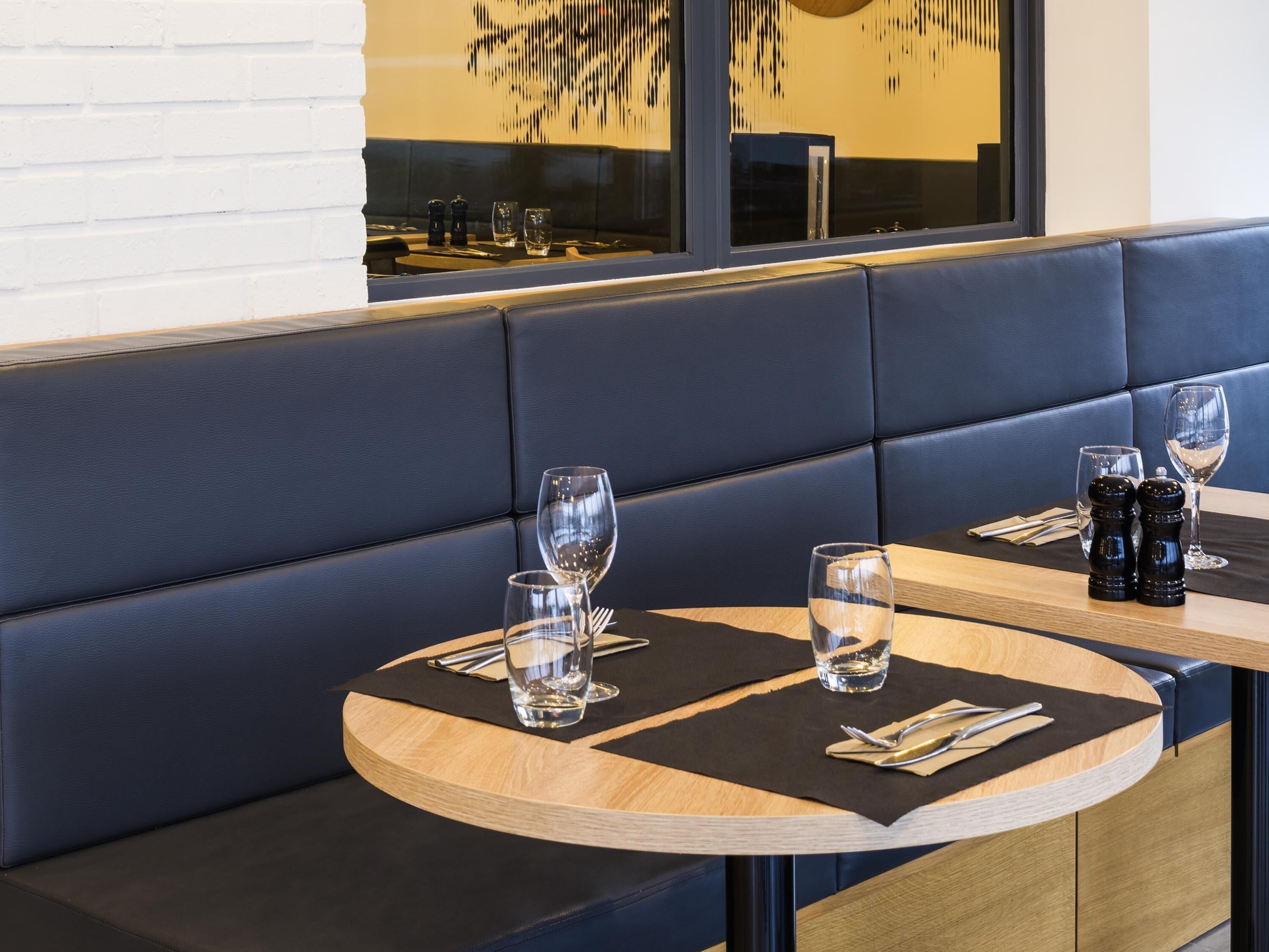 Interior de restaurante com assentos estofados em azul escuro, mesas redondas de madeira e jogos americanos pretos.