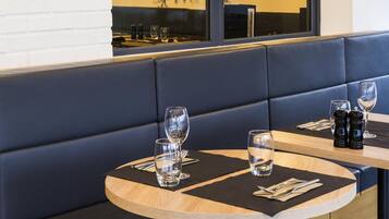 Interior de restaurante com assentos estofados em azul escuro, mesas redondas de madeira e jogos americanos pretos.