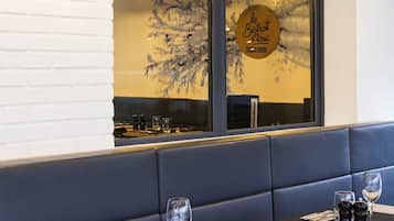 Interior de restaurante com área de assentos em camarote de couro escuro, uma mesa de madeira com copos de vinho e saleiros, e uma parede com uma placa decorativa pendurada.