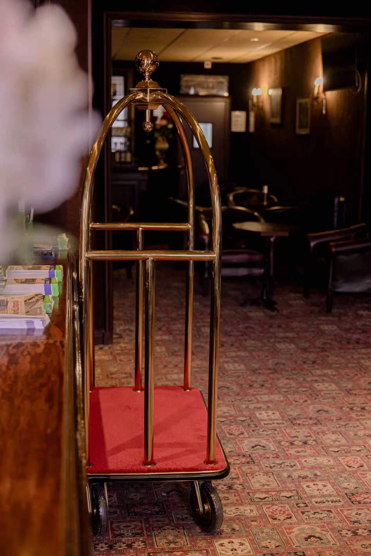 Quarto de hotel com carpete vermelho, carrinho dourado e uma pilha de notas de dinheiro sobre uma mesa.