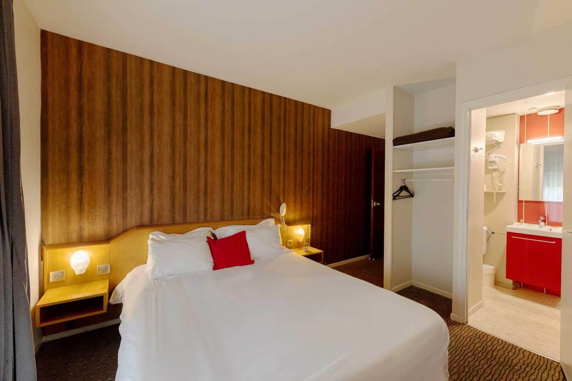 Quarto de hotel com cama, abajures, cabeceira de madeira, armário e banheiro com pia vermelha.