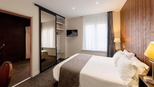 Quarto de hotel com cama, televisão, secretária com abajur, zona de chuveiro e guarda-roupa.