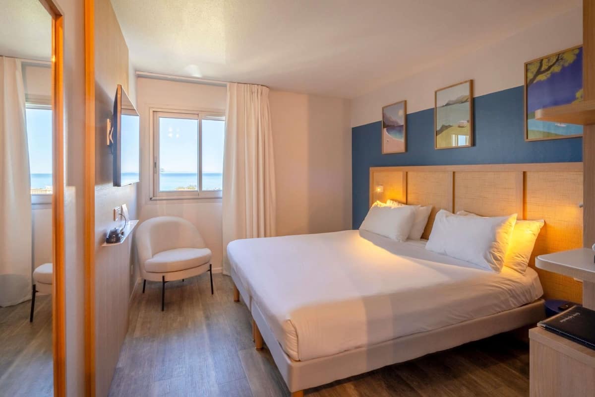 Quarto de hotel com uma cama grande, uma cadeira, uma escrivaninha e uma janela com cortinas.