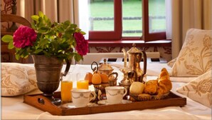 Bandeja de café da manhã com ovos, suco e doces sobre a cama, com vista para um jardim pela janela.