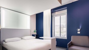 Quarto de hotel moderno com uma cama grande, uma pequena escrivaninha, uma cadeira e uma janela com vista para a cidade.