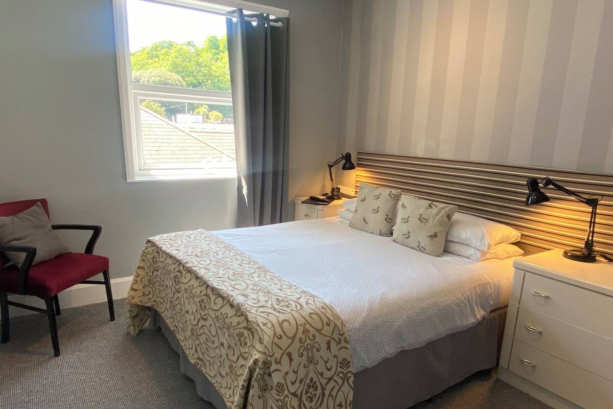 Quarto com cama, uma cadeira, mesa de cabeceira e janela com cortinas.
