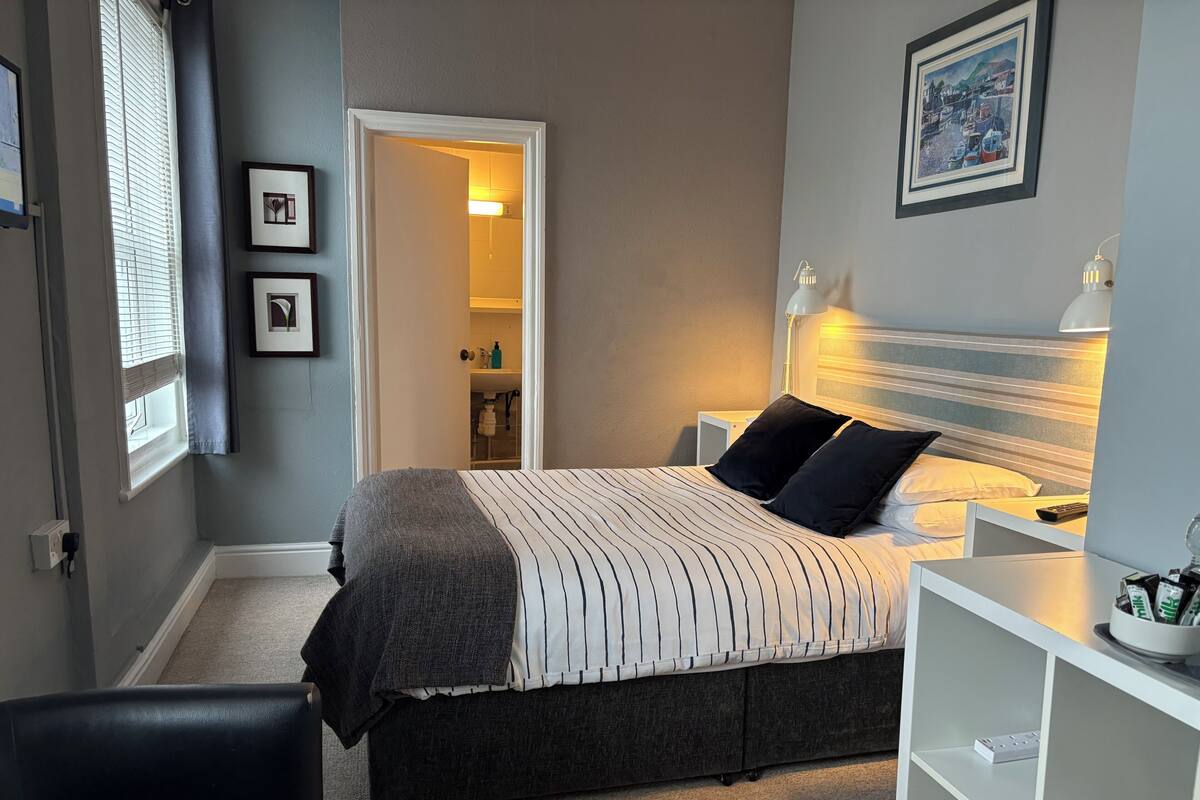 Um quarto com cama, mesas de cabeceira, uma cadeira, escrivaninha e um quadro na parede.