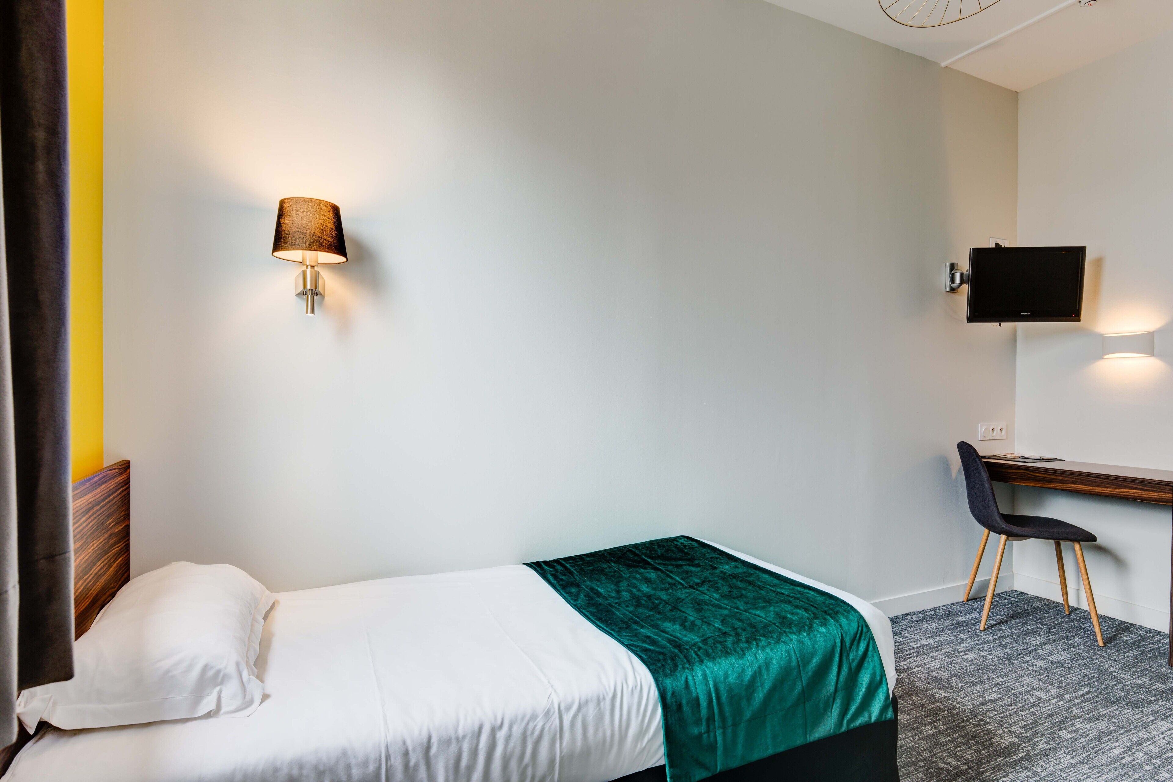 Quarto de hotel com cama, escrivaninha, cadeira, televisão e luminária fixada na parede.