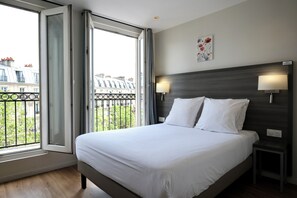 Quarto de hotel com uma cama grande, duas luminárias de cabeceira, uma janela com vista para edifícios e uma mesa pequena.