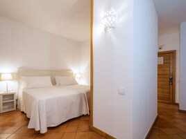 Quarto de hotel com cama, mesas de cabeceira com luminárias, porta de madeira e parede branca.