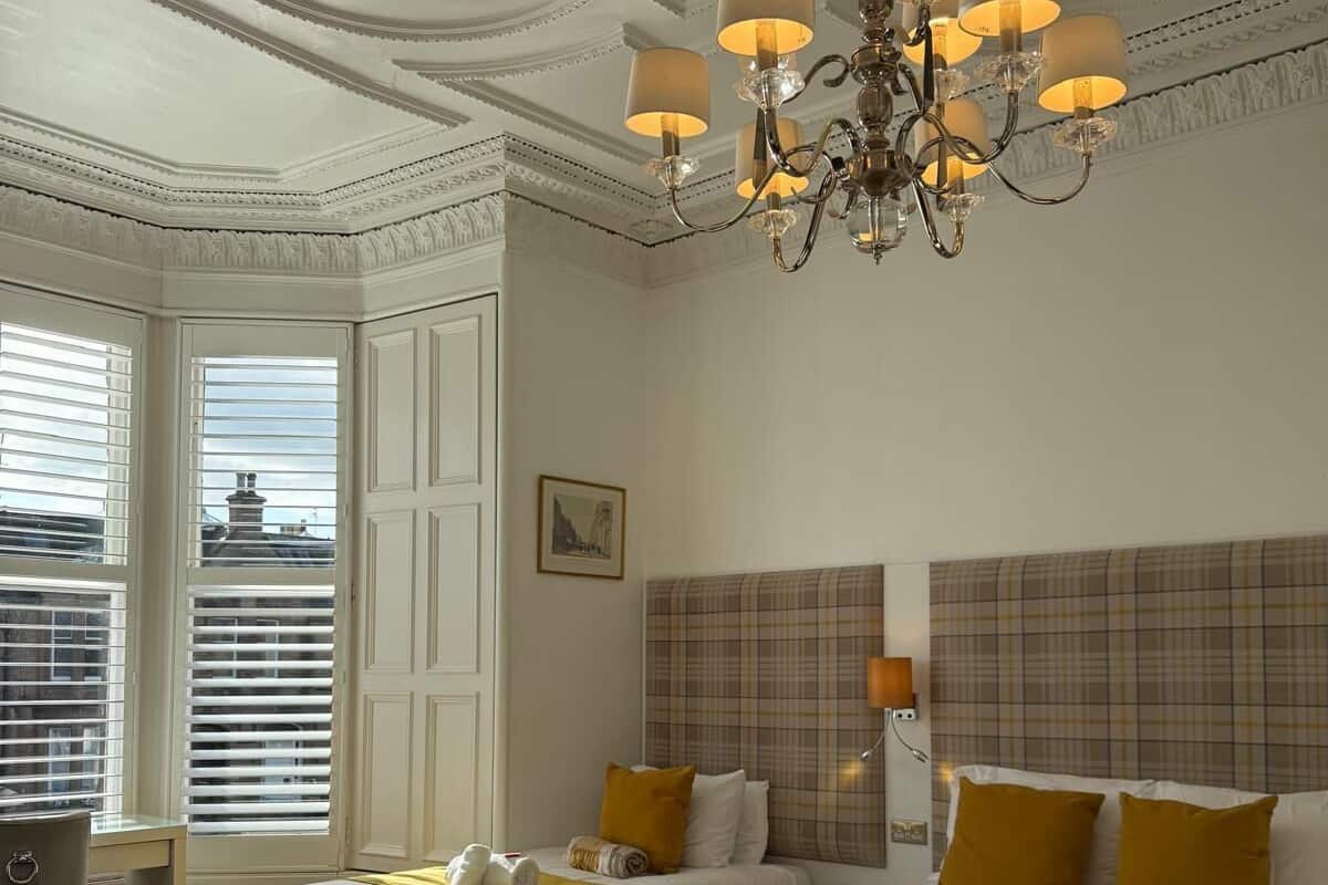 Quarto de hotel com duas camas, um candelabro, uma escrivaninha e uma janela com persianas.