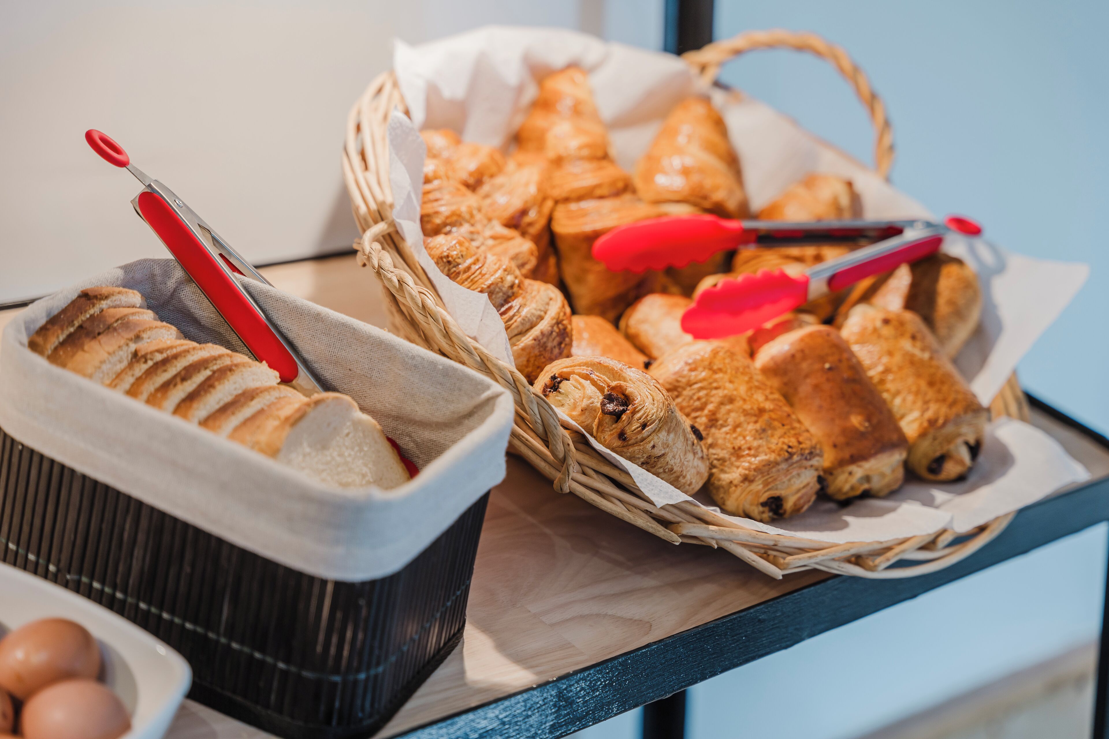 Uma bandeja com diversos pães e doces, incluindo croissants e baguetes, com pinças vermelhas por cima.