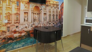 Quarto com um mural de um edifício imponente, uma televisão de tela plana na parede, uma mesa redonda com duas cadeiras e um micro-ondas em um armário.
