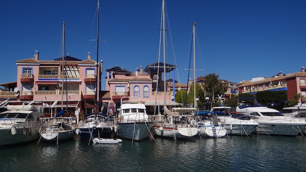 Uma marina com vários barcos atracados, rodeada por edifícios coloridos com varandas e um céu azul límpido.
