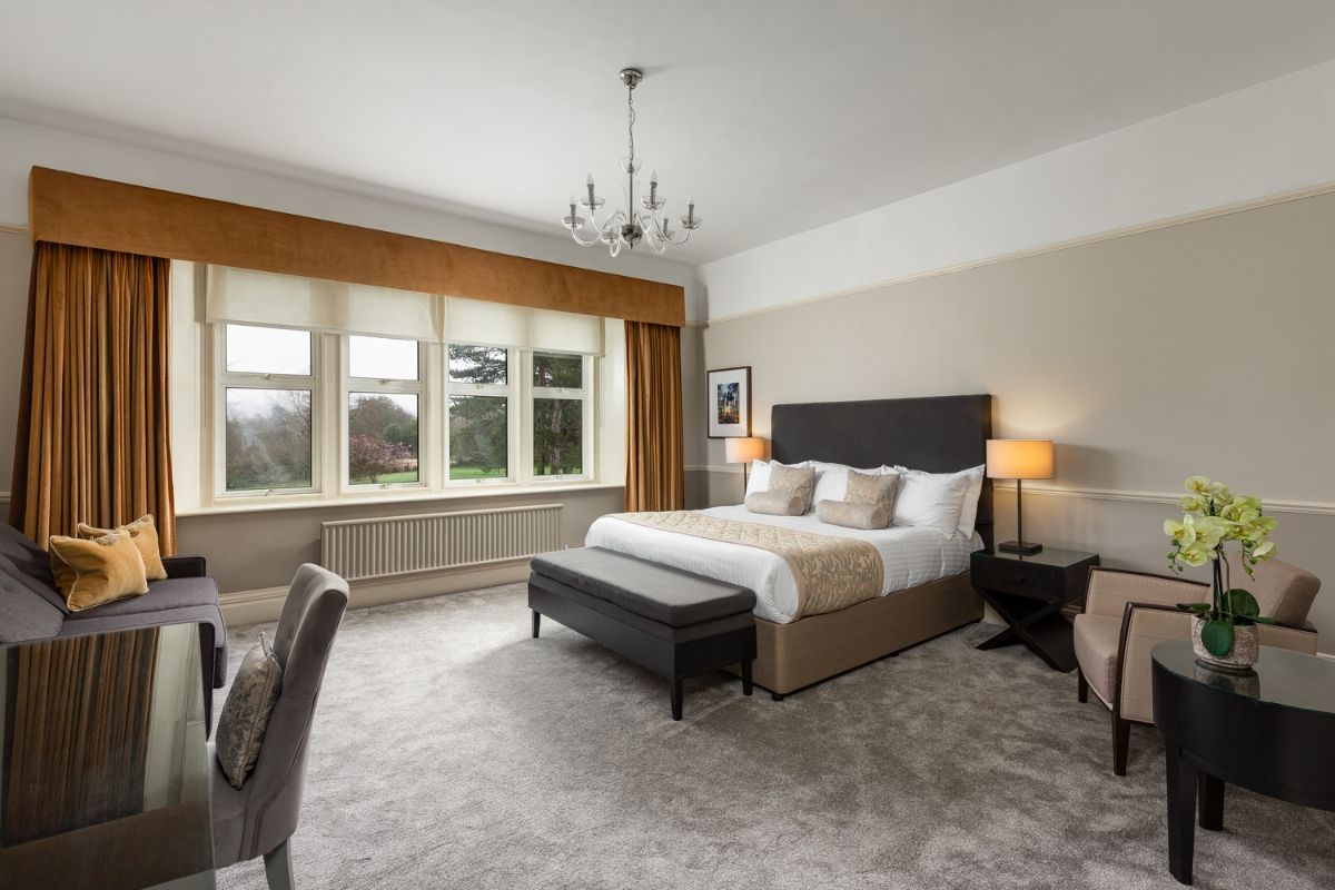 Quarto de hotel com uma cama grande, escrivaninha, cadeira, banqueta e vista para o exterior pela janela.