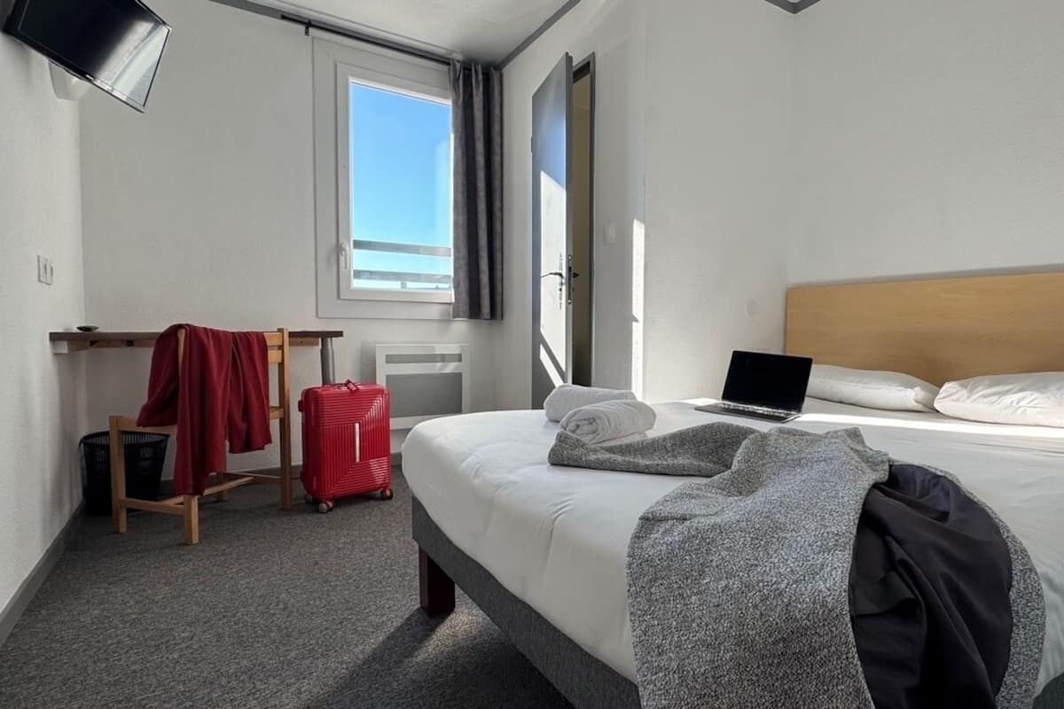 Quarto de hotel com uma cama, um laptop, uma televisão, uma janela e uma mala vermelha.