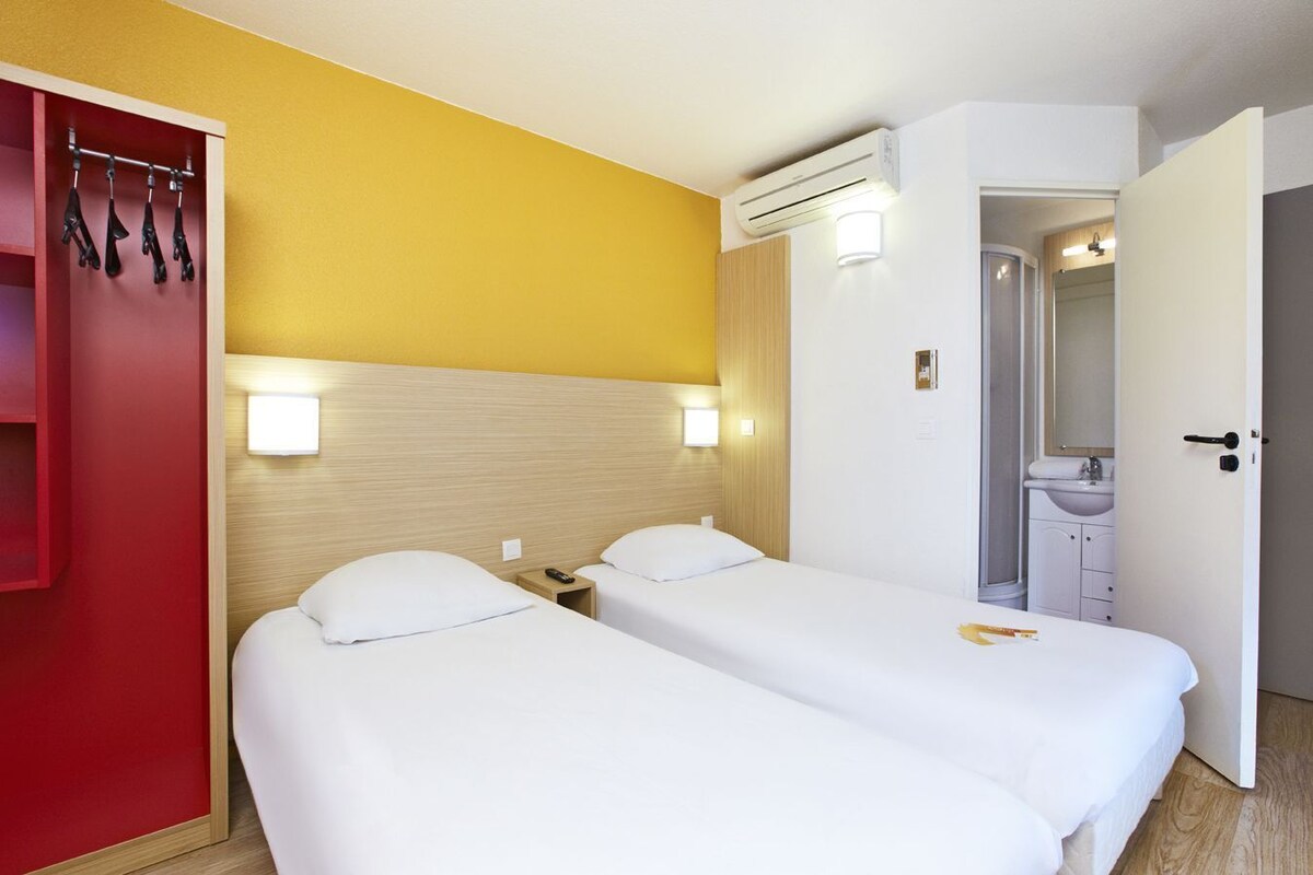 Quarto de hotel com duas camas, um guarda-roupa vermelho, banheiro com pia e espelho, e aparelho de ar condicionado.