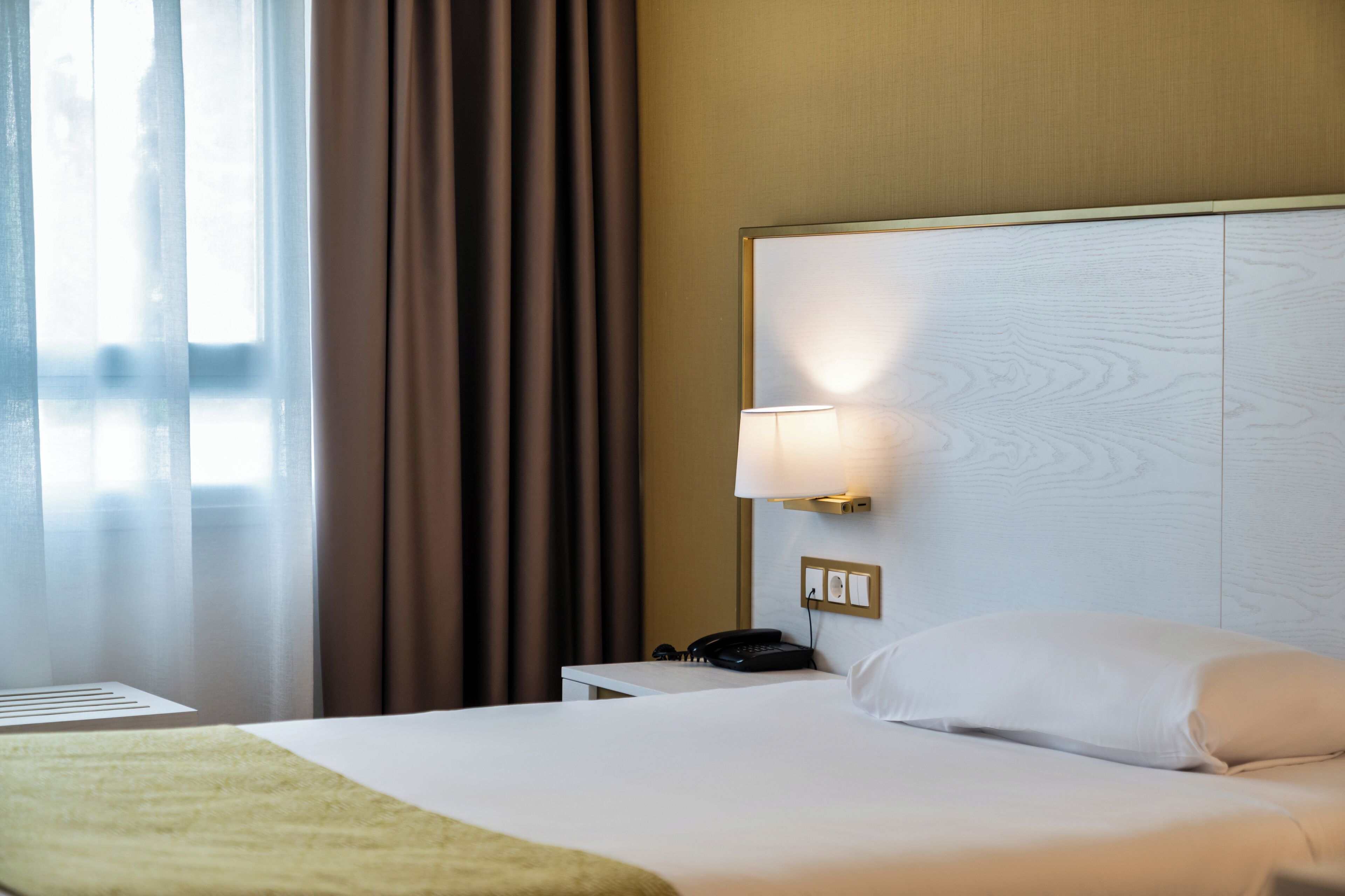 Quarto de hotel com uma cama, abajur, telefone e janela com cortinas.