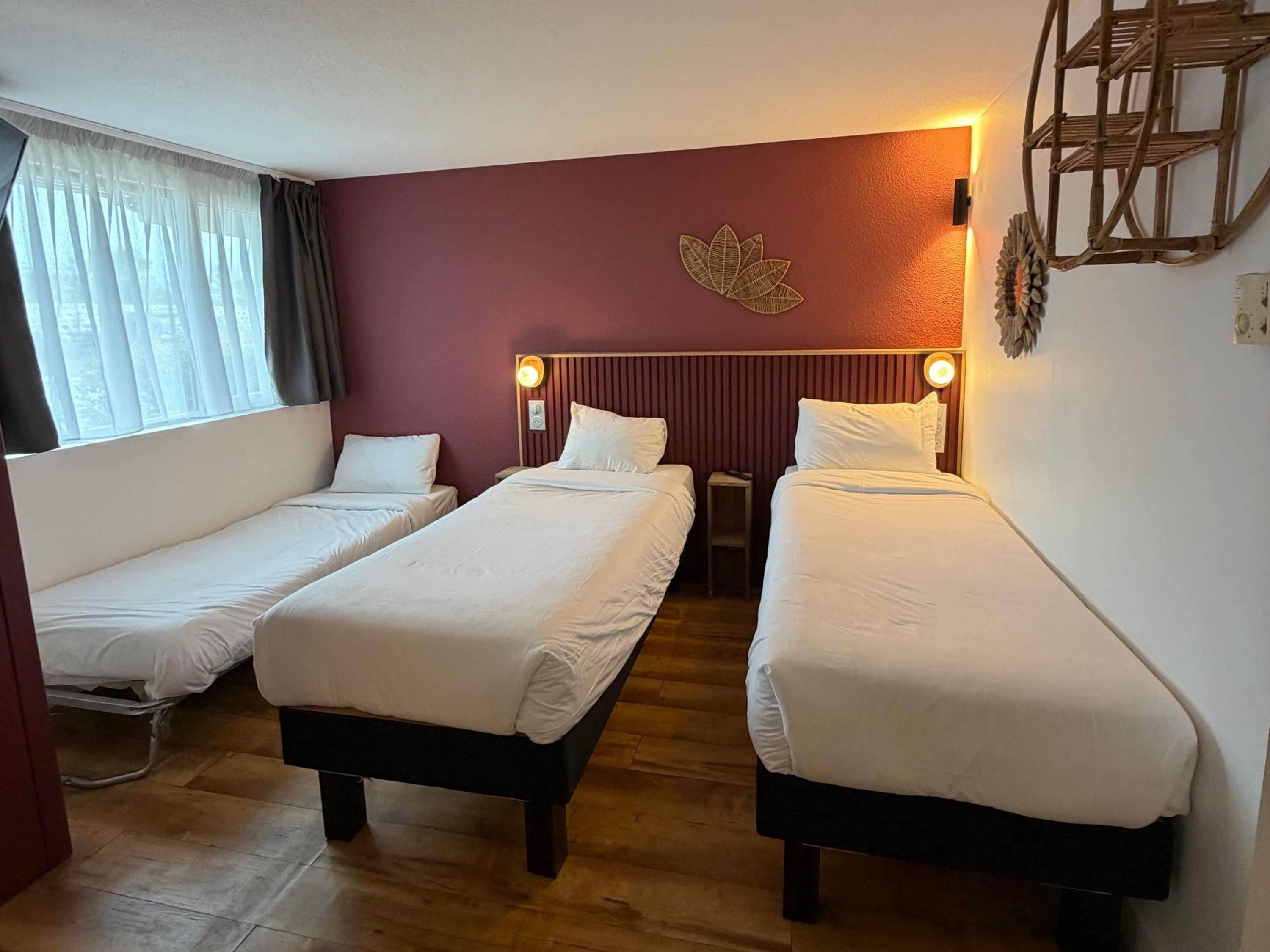 Quarto de hotel com duas camas de solteiro, janela com cortinas, luminária na parede e um objeto decorativo na parede.
