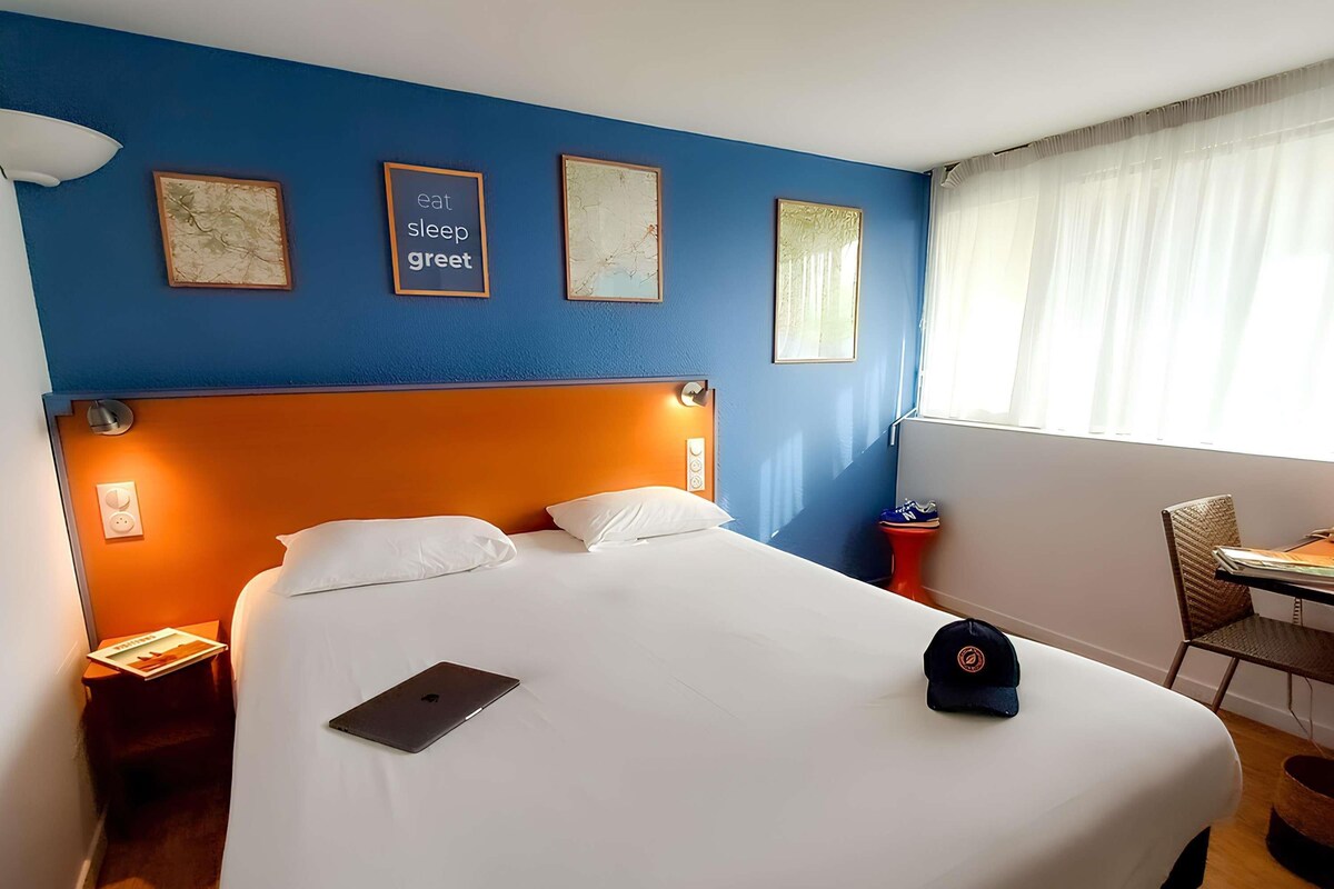 Quarto de hotel com uma cama grande, uma mesa e uma cadeira. Há três quadros na parede, um abajur e uma janela com cortinas.