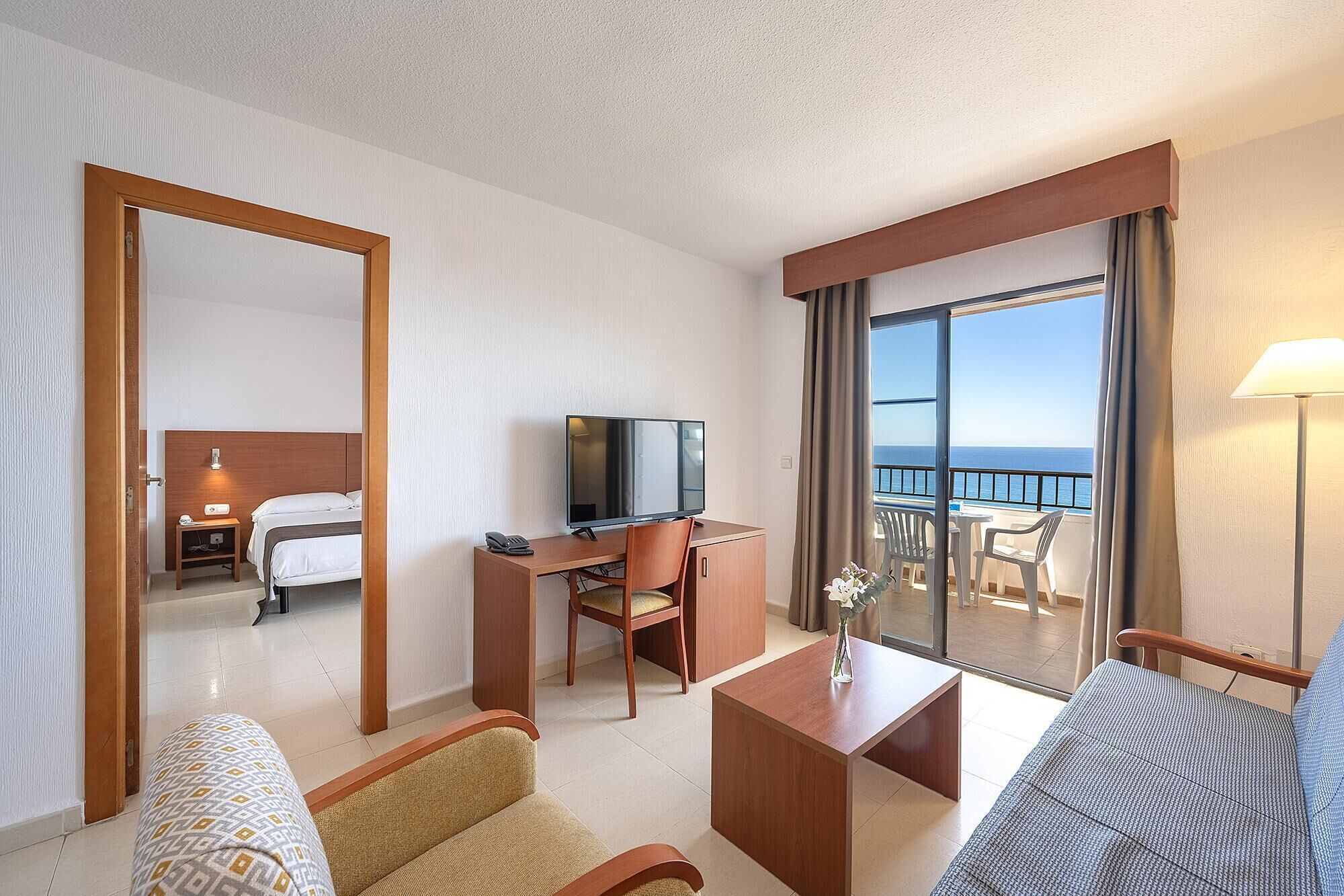 Quarto de hotel com cama, mesa de trabalho com televisão, varanda com vista para o mar e área de estar com luminária.