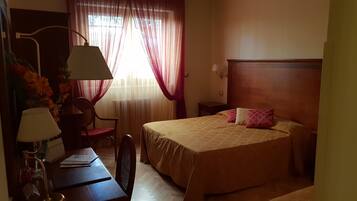 Quarto de hotel com cama, escrivaninha com abajur, cadeira e janela com cortinas.