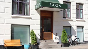 Entrada de hotel com toldo verde identificado como "SAGA HOTEL", um banco de madeira, duas plantas em vasos e uma bicicleta estacionada na frente.
