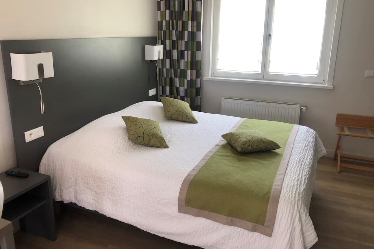 Cama bem arrumada com colcha branca e verde, duas almofadas combinando, mesa de cabeceira com abajur, janela com cortinas e radiador.