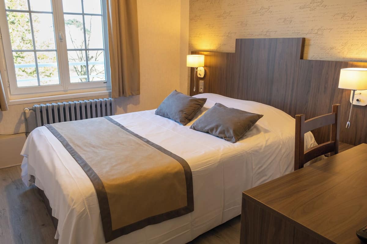 Cama bem arrumada com cabeceira, duas luminárias de apoio, mesa de cabeceira de madeira e uma janela com cortinas.