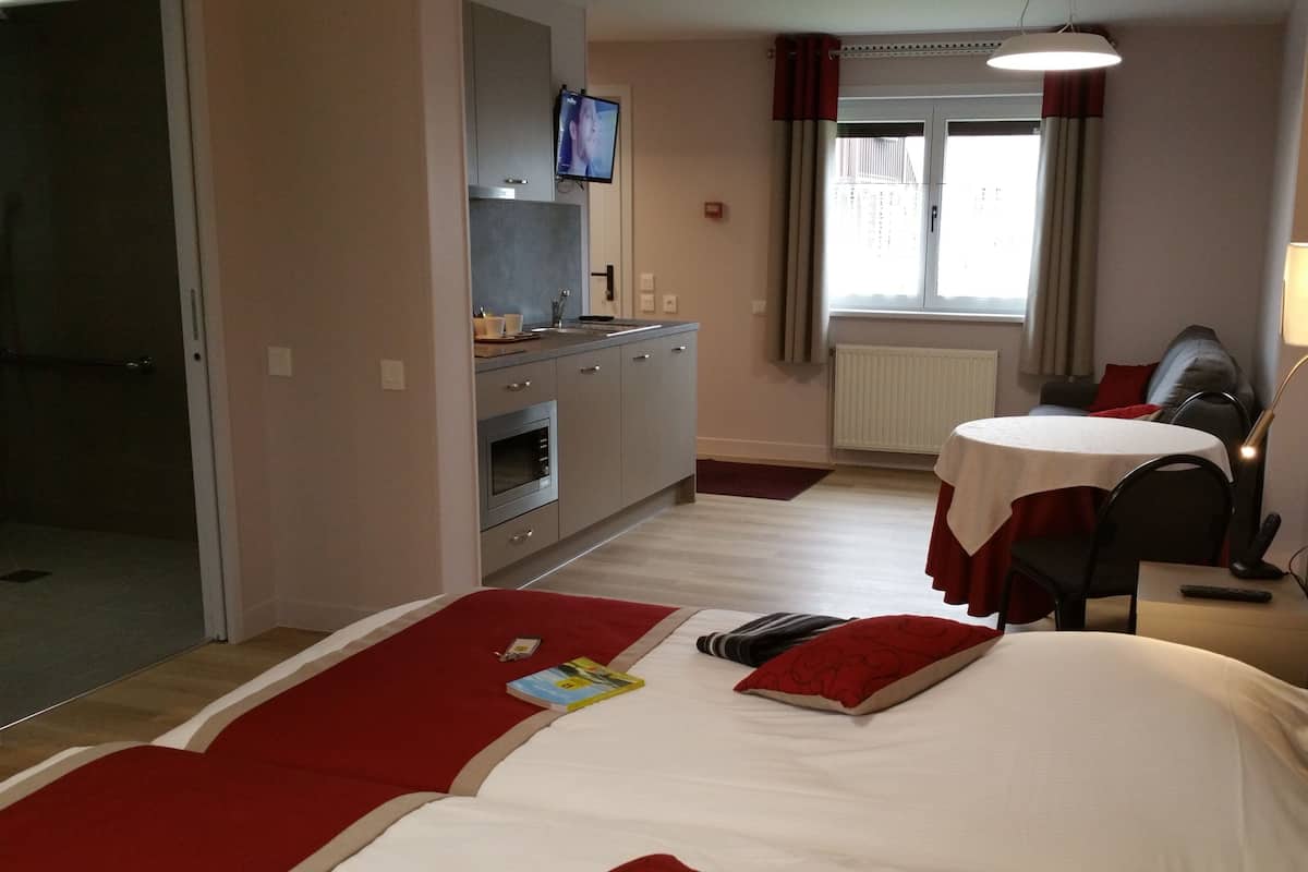Quarto de hotel com cama, escrivaninha, cadeira, televisão, janela com cortinas, radiador e cozinha compacta.
