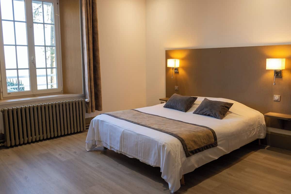 Quarto de hotel com uma cama grande, duas luminárias de cabeceira, uma janela com cortinas e um radiador.