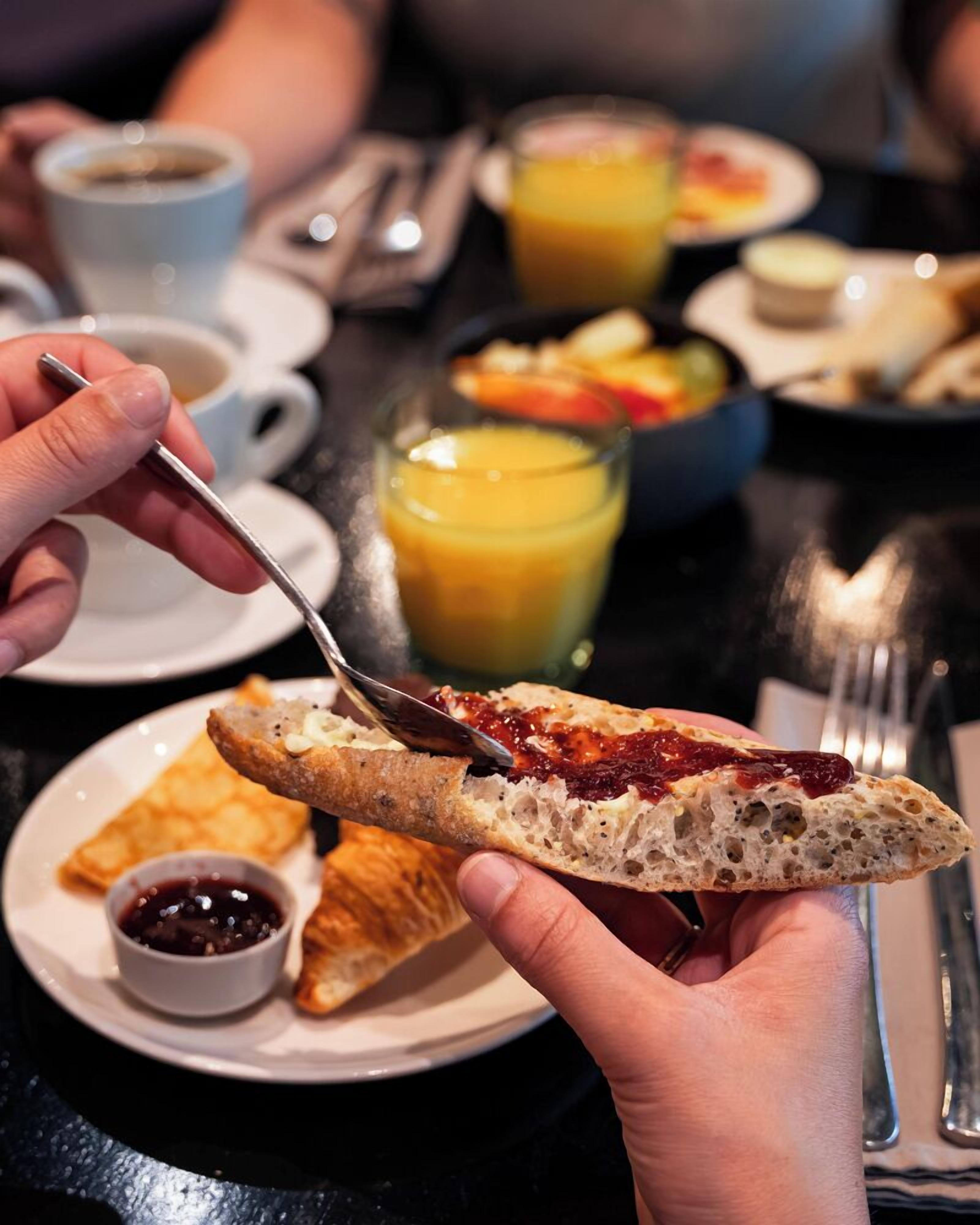 Uma mesa posta para o café da manhã, com uma pessoa espalhando geleia no pão, uma xícara de café e um copo de suco de laranja.