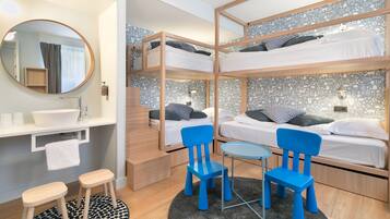 Quarto de hotel compacto com beliches, um espelho redondo, uma mesa pequena e duas cadeiras azuis.