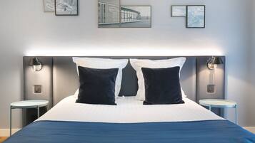 Uma cama bem arrumada com um cobre-leito branco e azul, duas almofadas pretas e duas mesas de cabeceira com luminárias.
