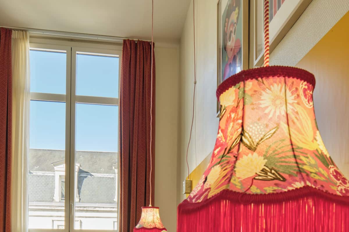 Quarto de hotel com uma cama, janela com cortinas, luminária e obras de arte na parede.