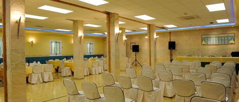 Uma sala de banquetes com fileiras de cadeiras, um palco com um pódio e uma área de buffet.