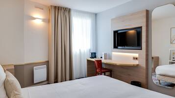 Quarto de hotel com uma cama, uma mesa com cadeira, TV fixada na parede, um espelho e uma janela com cortinas.