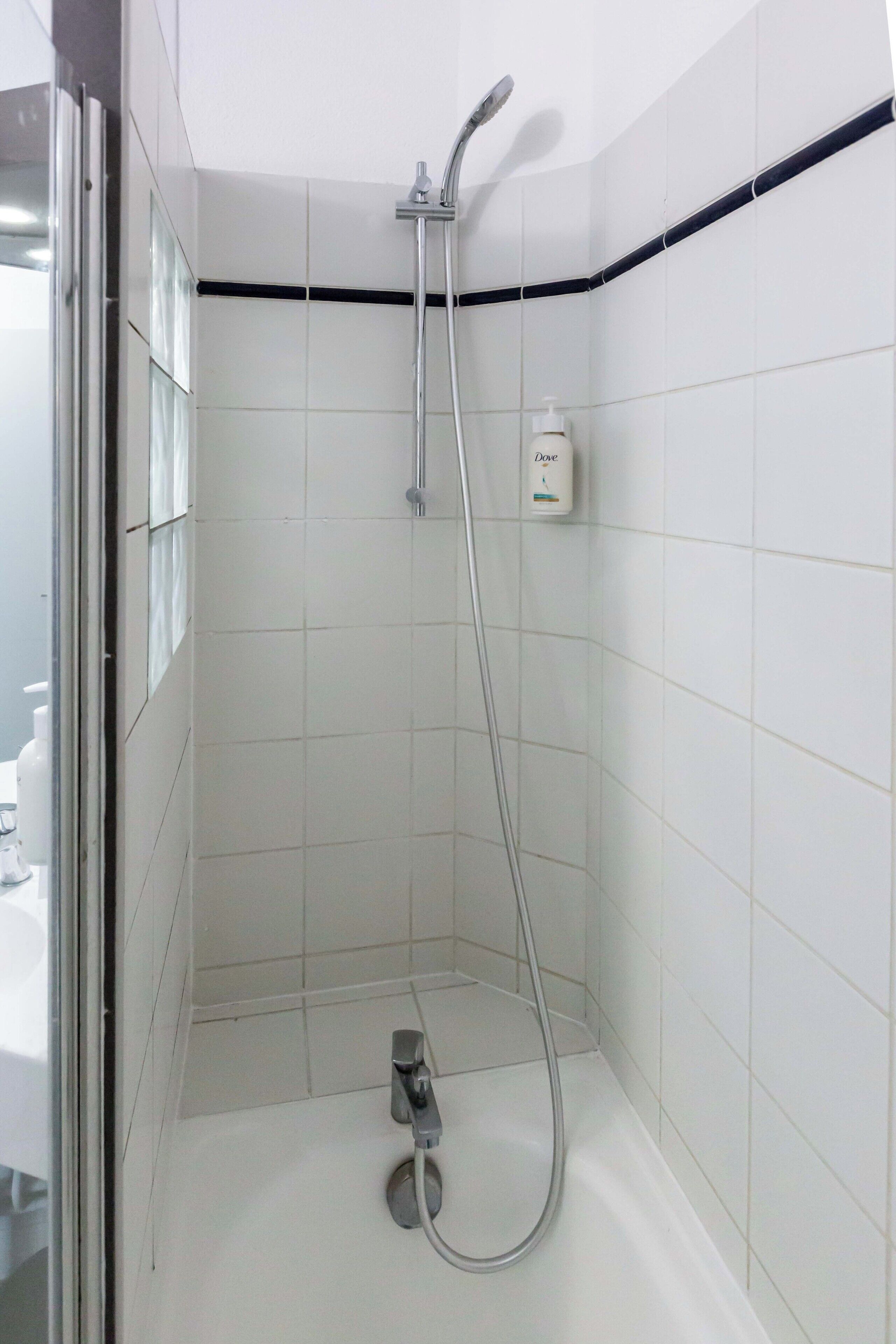Chuveiro com chuveiro manual e torneira fixada na parede, um sabonete Dove e uma porta de vidro.