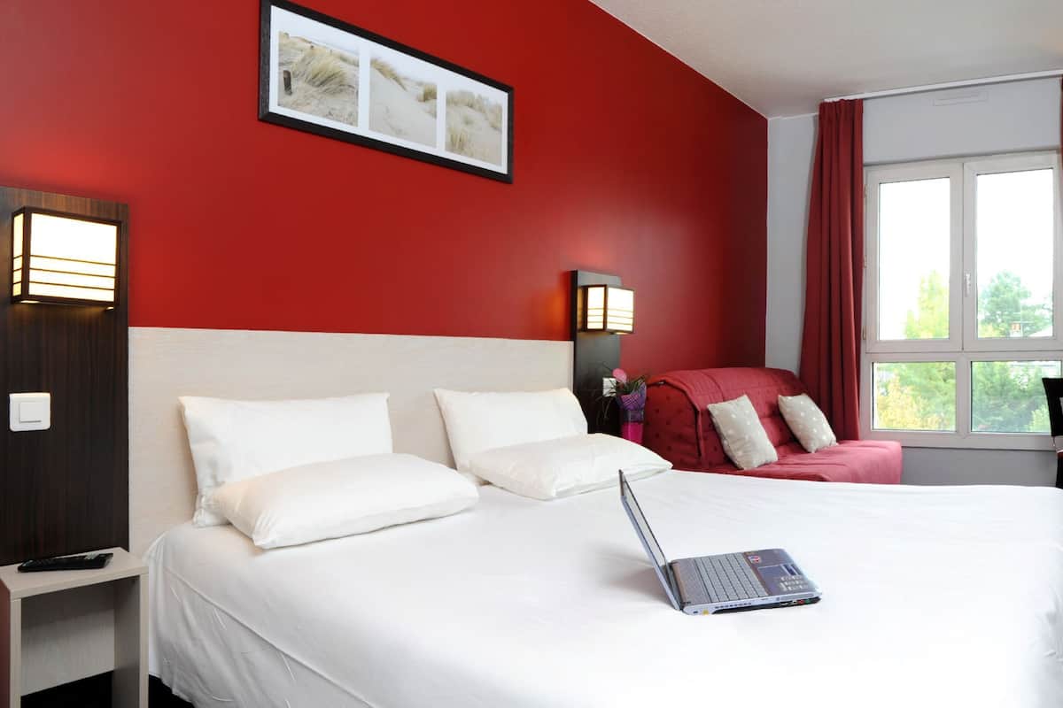 Quarto de hotel com uma cama grande, uma escrivaninha, uma cadeira, uma televisão e uma janela com cortinas.